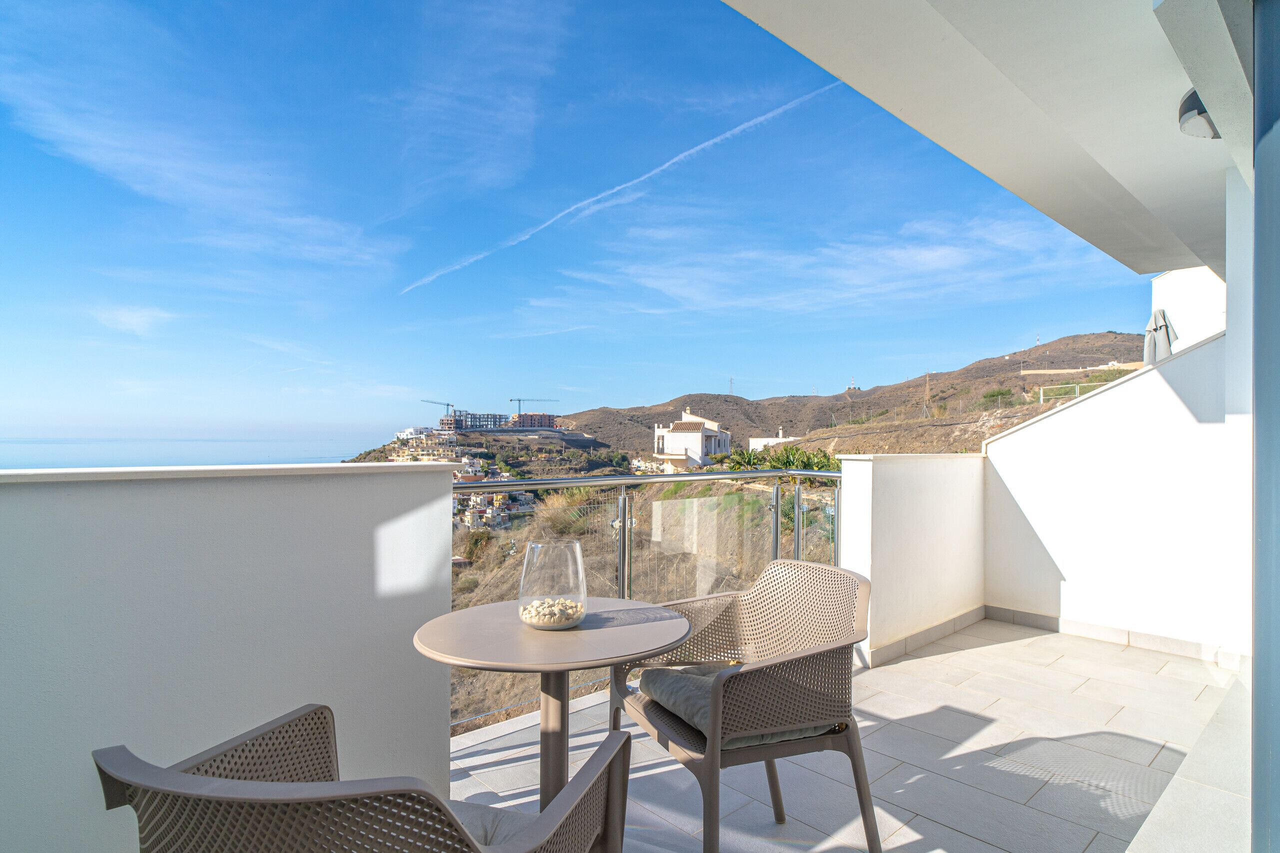 Varanda com mesa e duas cadeiras, vista para paisagem costeira com edifícios e colinas.