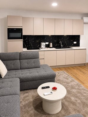 Uma sala de estar moderna com um sofá seccional cinza, uma mesa de centro redonda e uma área de cozinha com revestimento de mármore preto.
