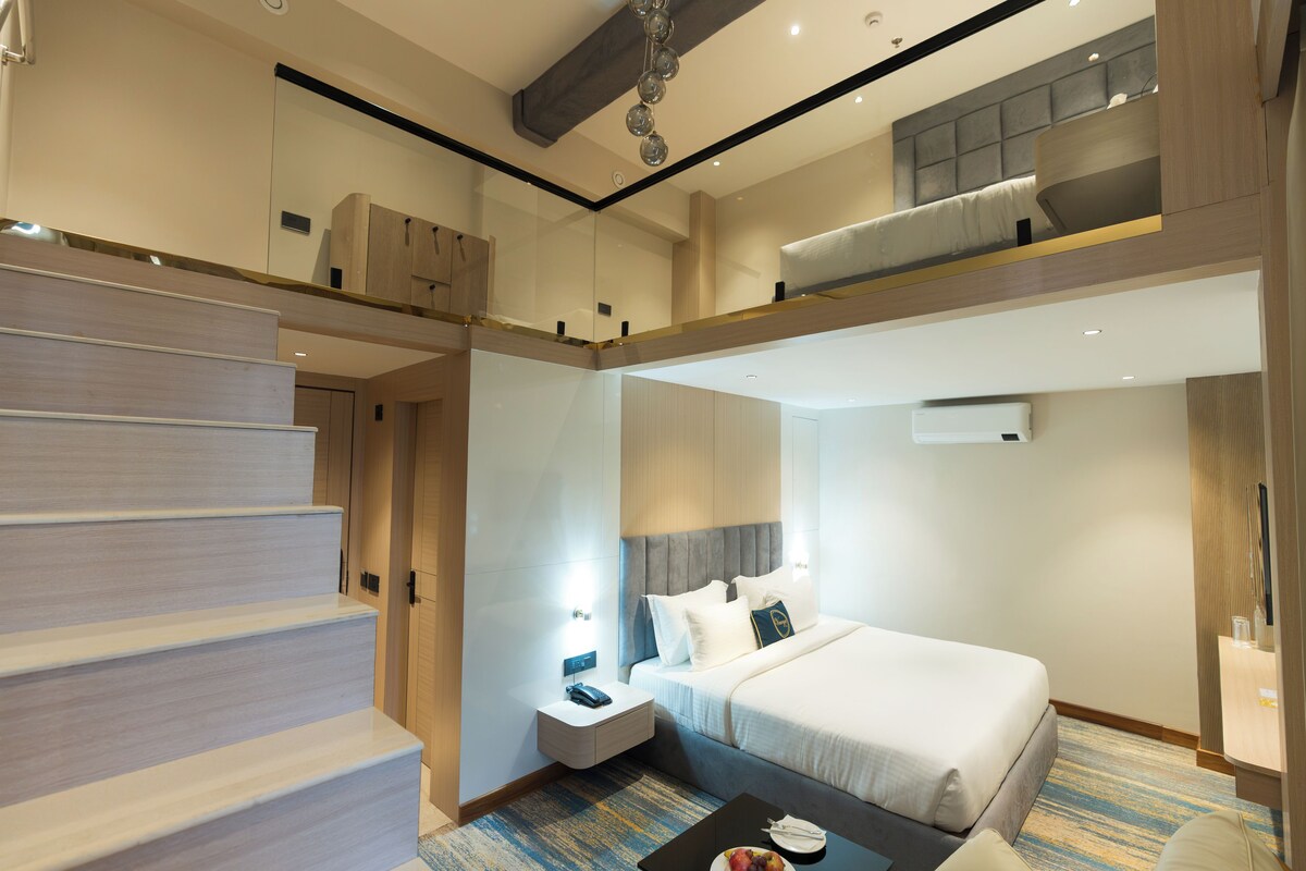 Quarto de hotel com cama, escrivaninha, telefone, mesa pequena com uma tábua de frutas e escada.