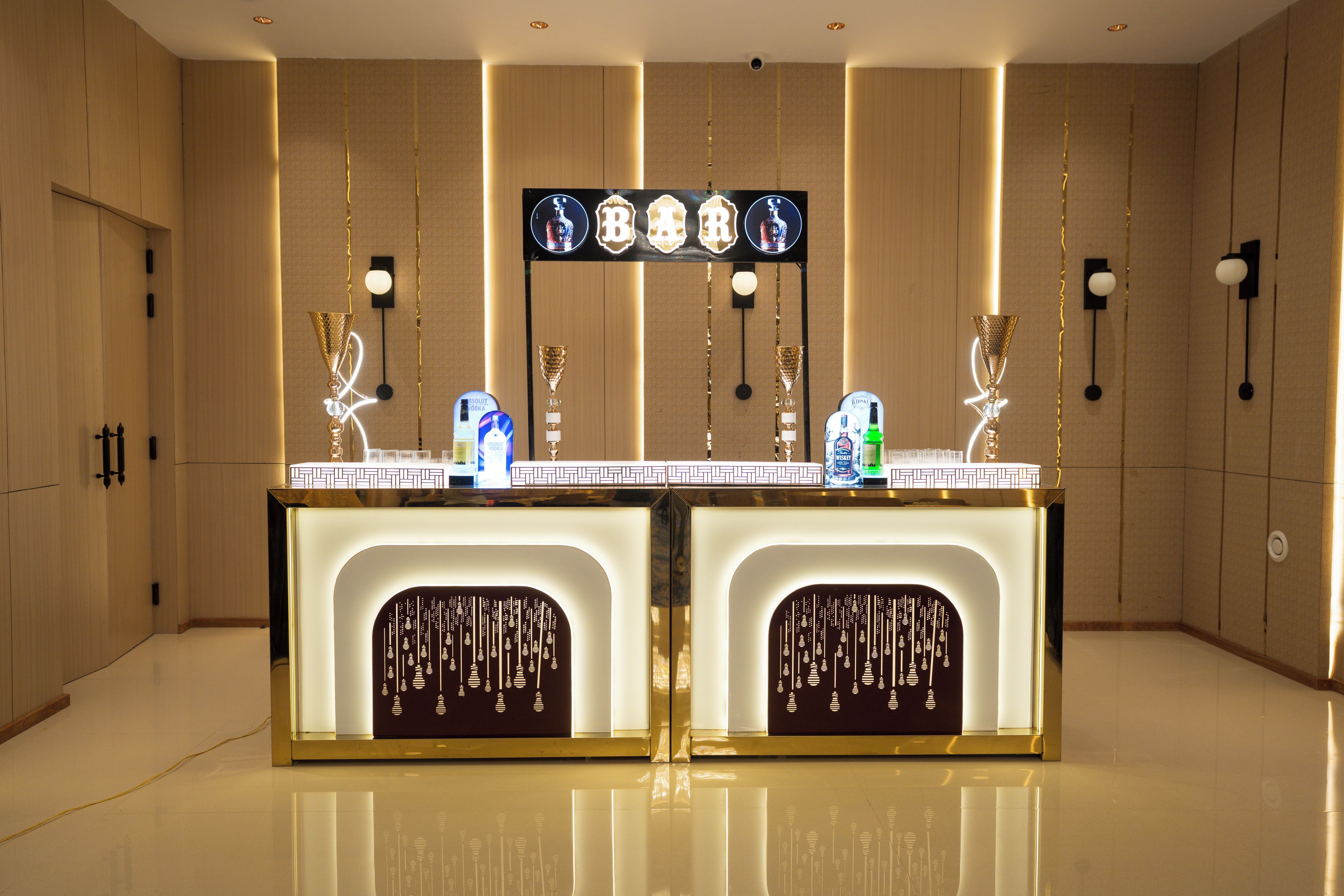 Uma área de bar com dois balcões, luminárias fixadas na parede e uma tela grande exibindo um logotipo.