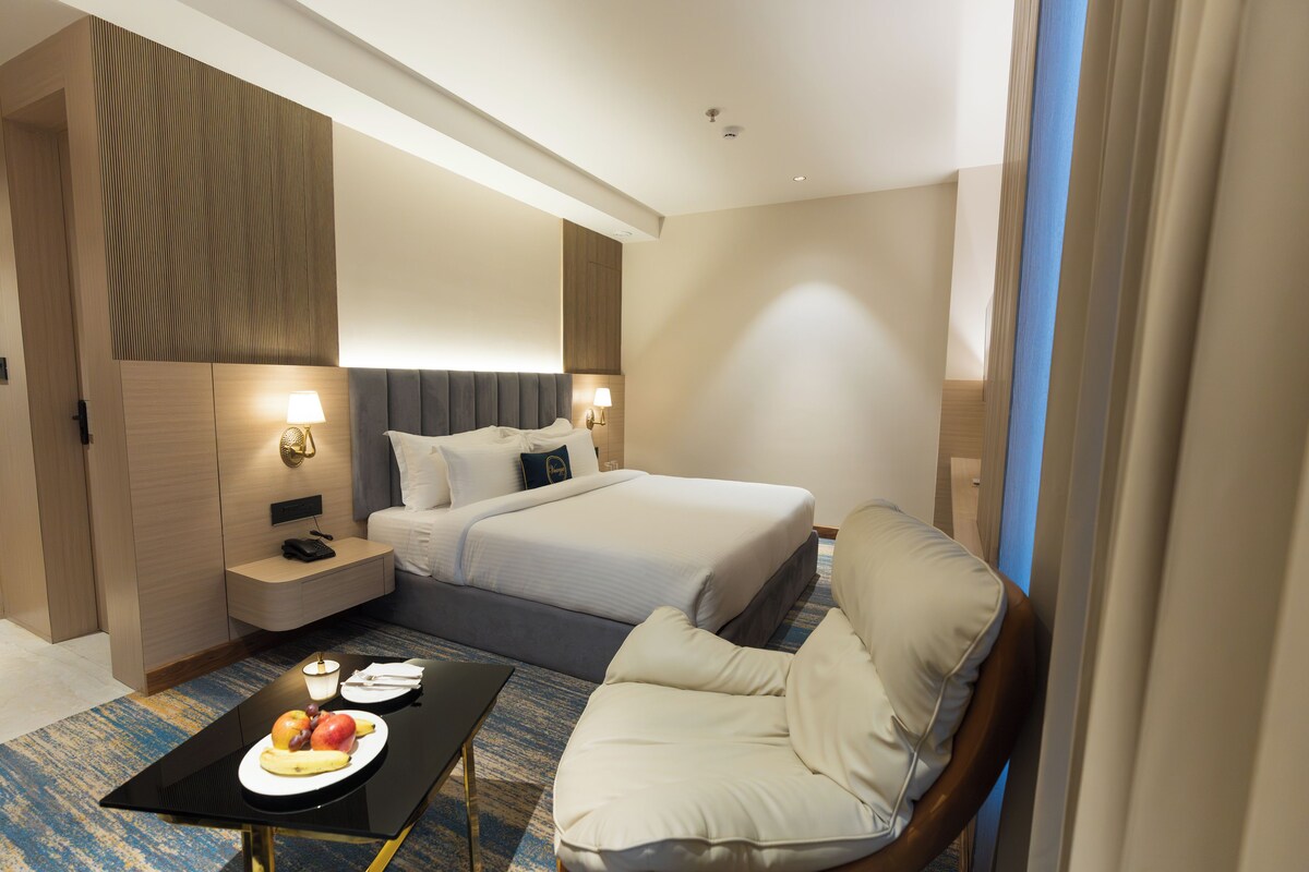 Quarto de hotel com uma cama, uma escrivaninha com uma cesta de frutas, uma cadeira e uma mesa de cabeceira com abajur.