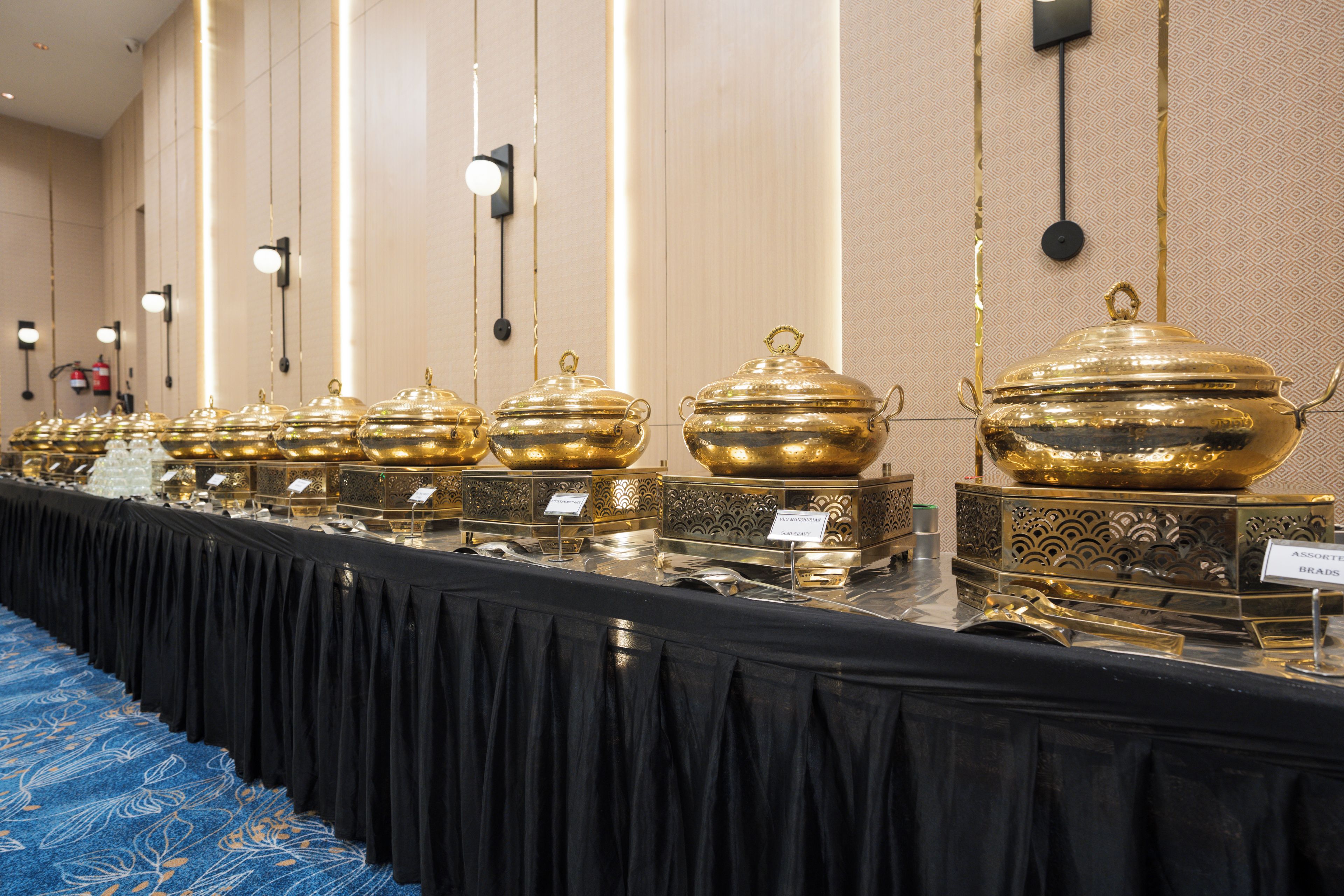 Uma mesa comprida com vários utensílios dourados para servir e recipientes, provavelmente para um buffet ou serviço de catering.