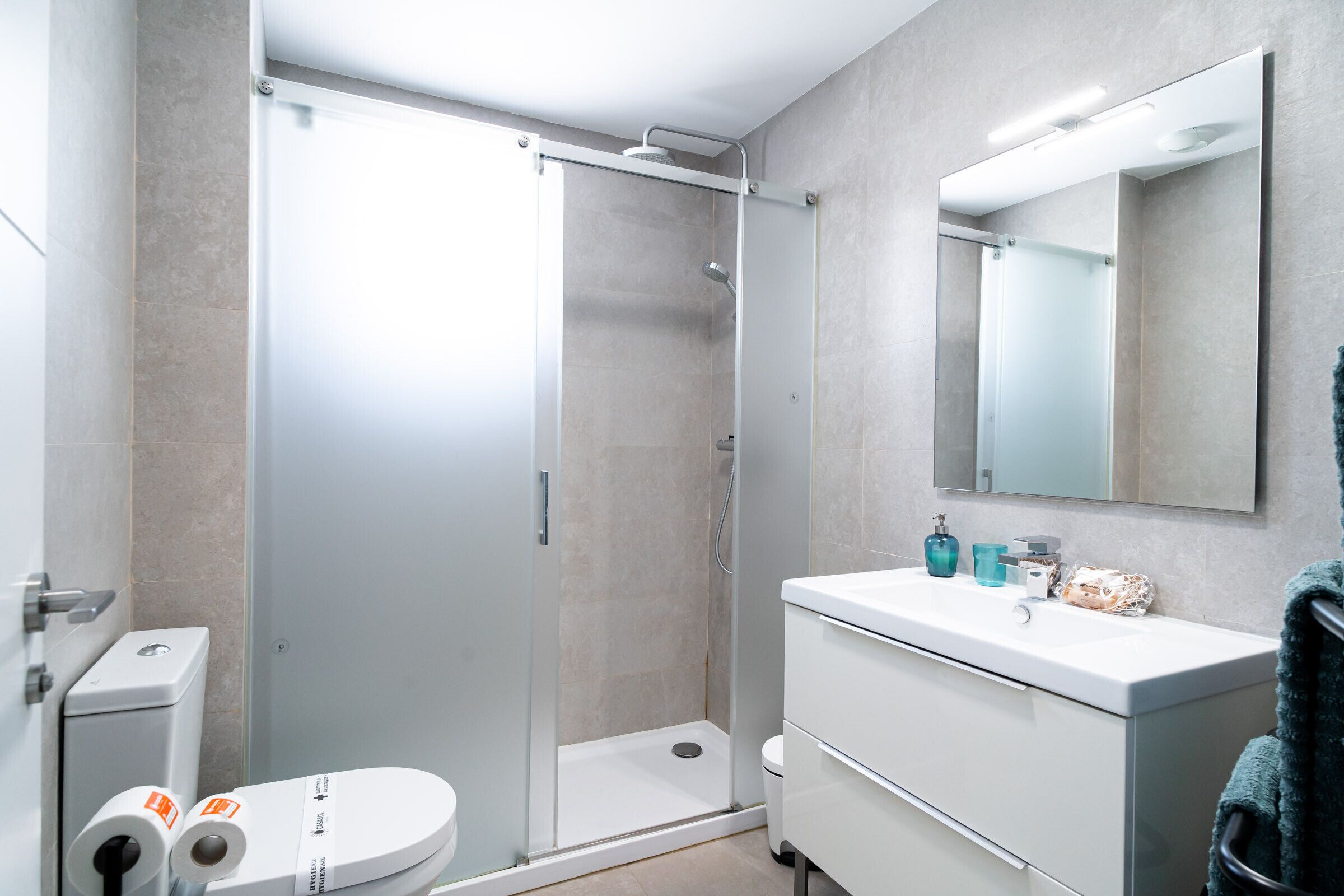 Uma casa de banho moderna com cabine de duche em vidro, lavatório branco com artigos de higiene pessoal e espelho sobre o lavatório.