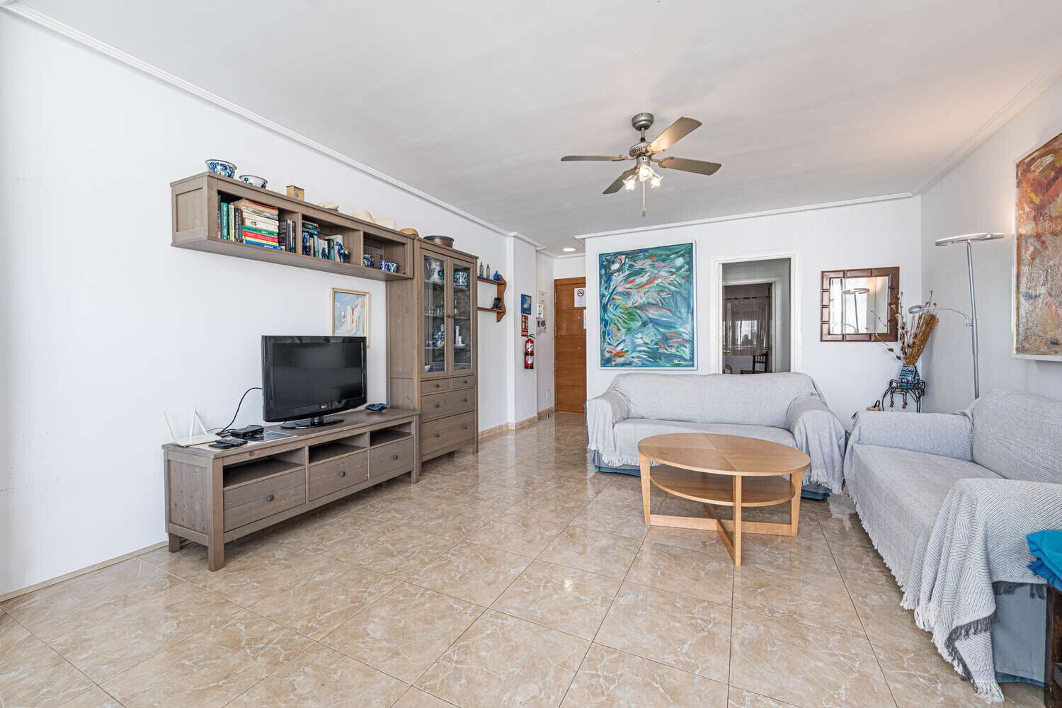 Sala de estar com televisão, painel de entretenimento em madeira, ventilador de teto, sofá e um quadro na parede.