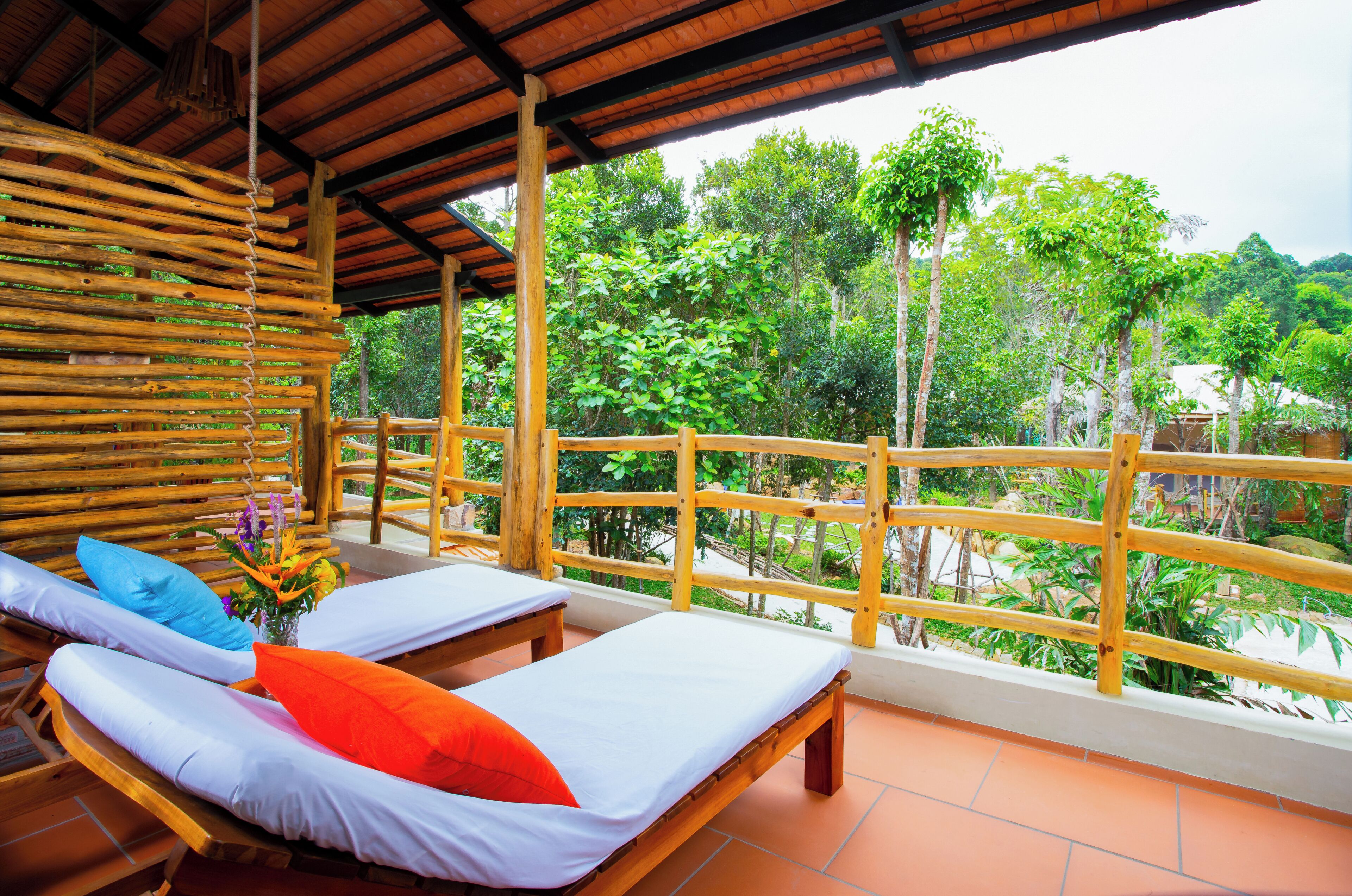 A wooden balcony with two beds, a hanging chair, and a view of lush greenery.