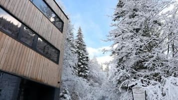 Uma trilha coberta de neve que leva a um edifício com design moderno, rodeado por árvores carregadas de neve.