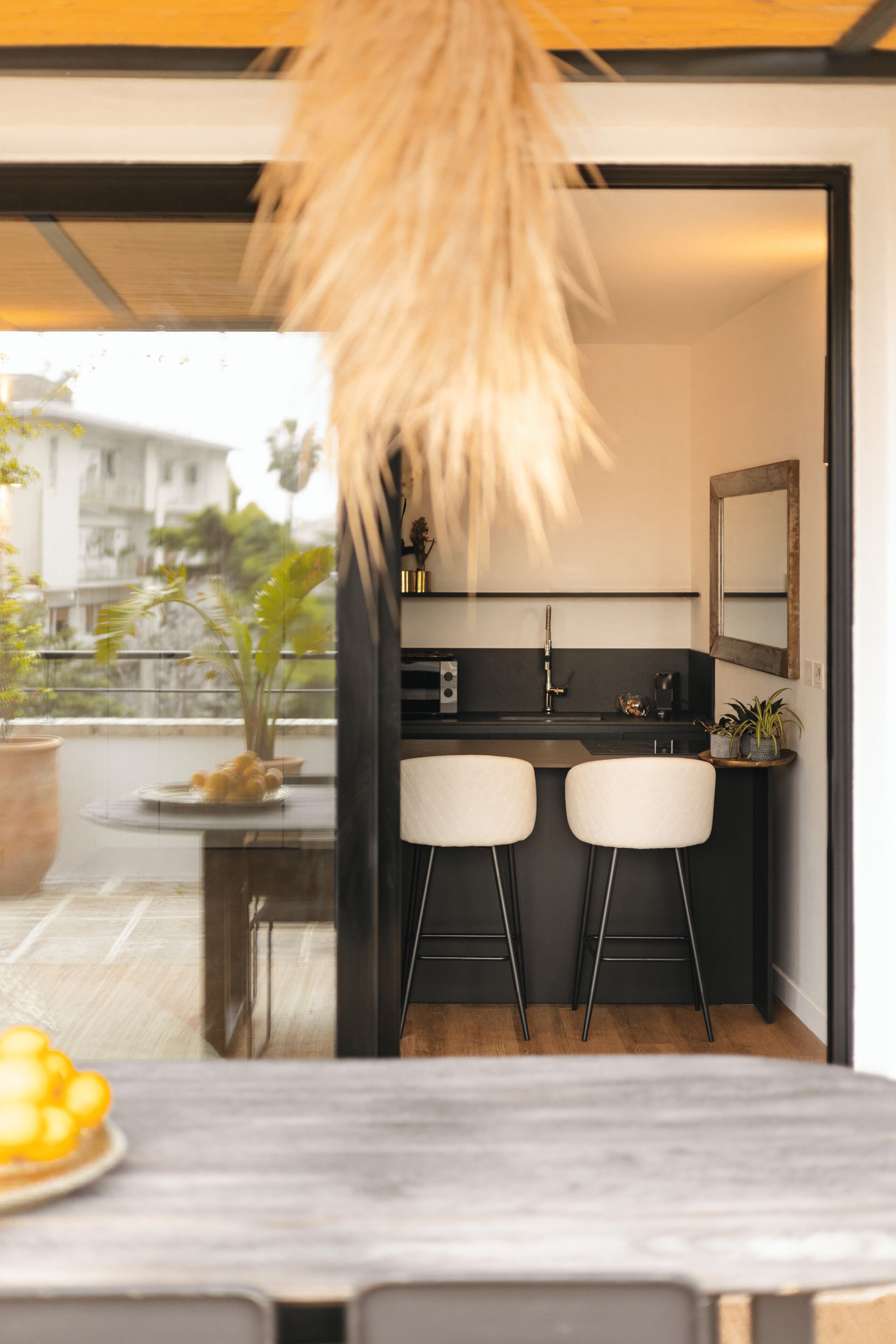 Uma cozinha moderna com bancada preta, dois banquetas brancas, luminária pendente e vista para a rua.