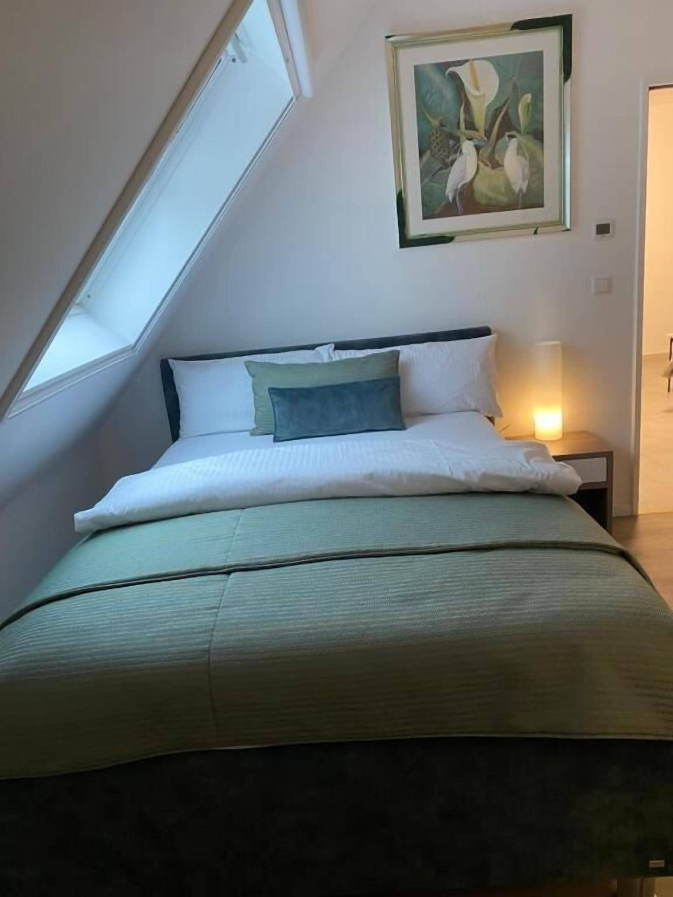 Quarto com cama, mesa de cabeceira, luminária na parede e claraboia.