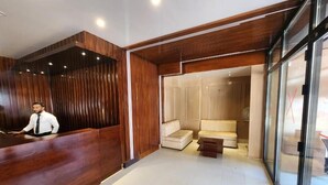 Uma área de recepção com balcão de madeira, sala de estar com sofás e divisória de vidro.