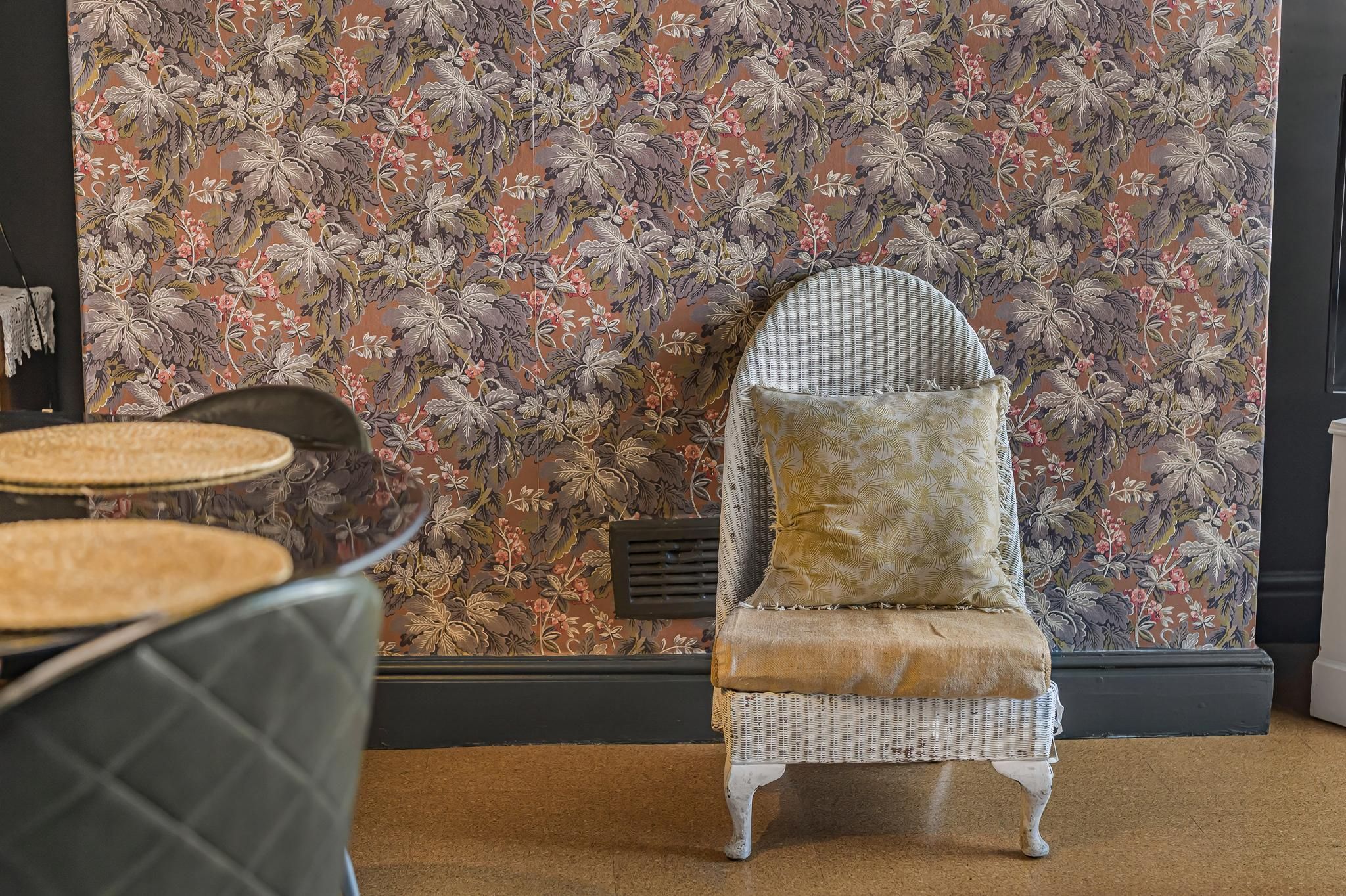 Uma cadeira de vime com almofada, papel de parede estampado com flores e uma ventilação na parede.