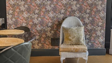 Uma cadeira de vime com almofada, papel de parede estampado com flores e uma ventilação na parede.