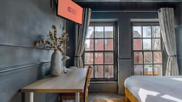Quarto com cama, escrivaninha com um vaso, televisão exibindo 'GUEST READY' e janela com cortinas.