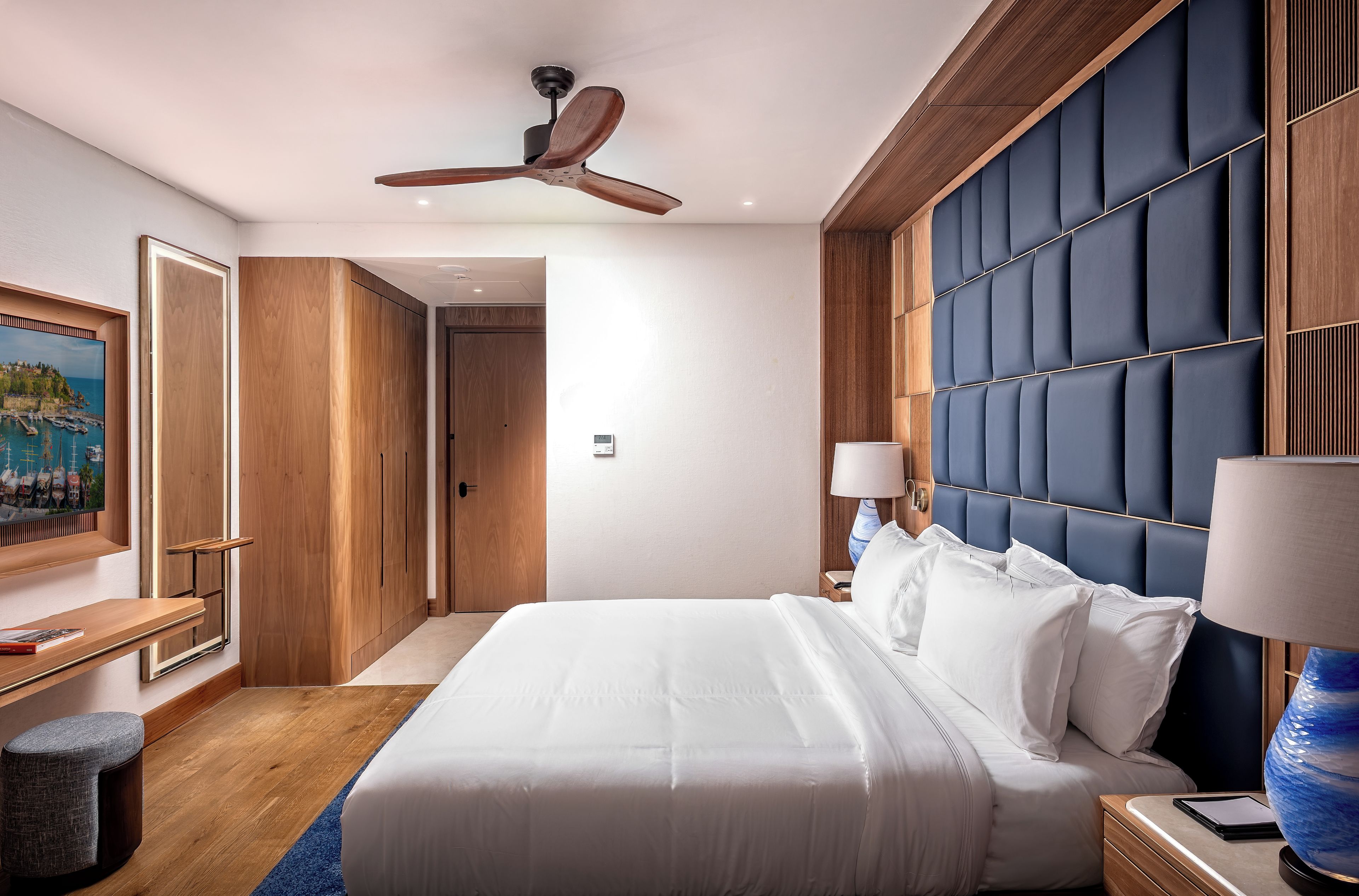 Quarto de hotel com uma cama grande, ventoinha no teto, guarda-roupa de madeira, mesa de cabeceira com luminária e um quadro na parede.