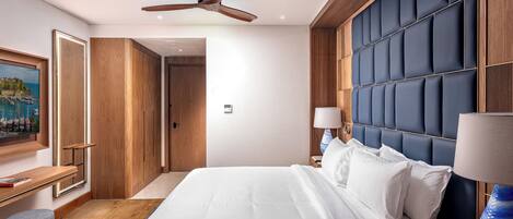Quarto de hotel com uma cama grande, ventoinha no teto, guarda-roupa de madeira, mesa de cabeceira com luminária e um quadro na parede.