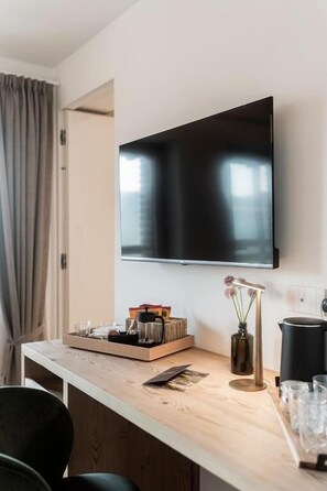 Quarto de hotel moderno com televisão de tela plana na parede, escrivaninha de madeira com um jogo de copos, um vaso com flores e uma cafeteira preta.