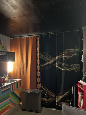 Quarto com cozinha compacta, parede colorida listrada, janela ampla e cortina com desenho de galho de árvore.