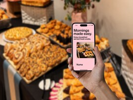 Mão segurando um smartphone exibindo um cardápio de café da manhã com diversos pães e doces.