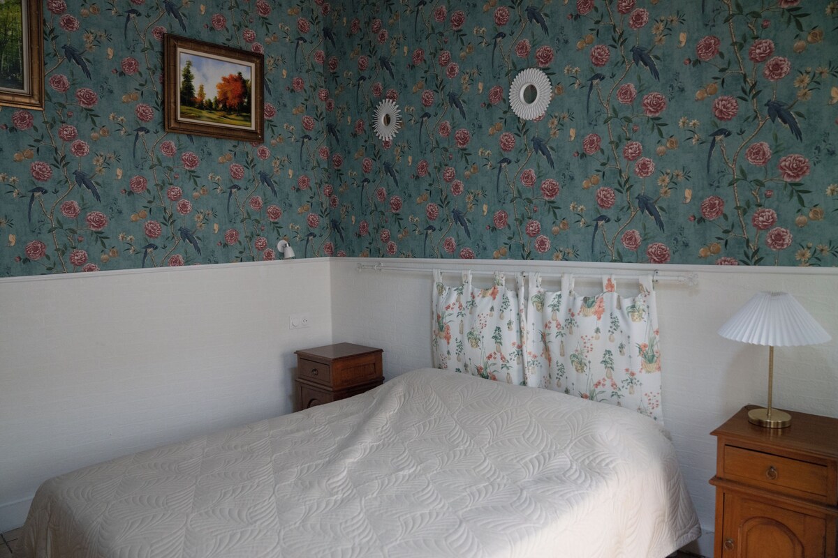Quarto com papel de parede floral, uma cama, mesa de cabeceira, abajur e um quadro na parede.