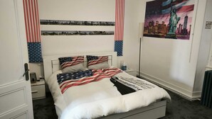 Quarto com cama, mesa de cabeceira, luminária e um quadro de uma paisagem urbana na parede.
