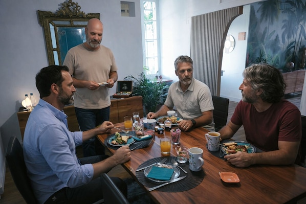 Um grupo de pessoas jantando em uma mesa de madeira em um quarto com um espelho grande, uma planta e um mural tropical.