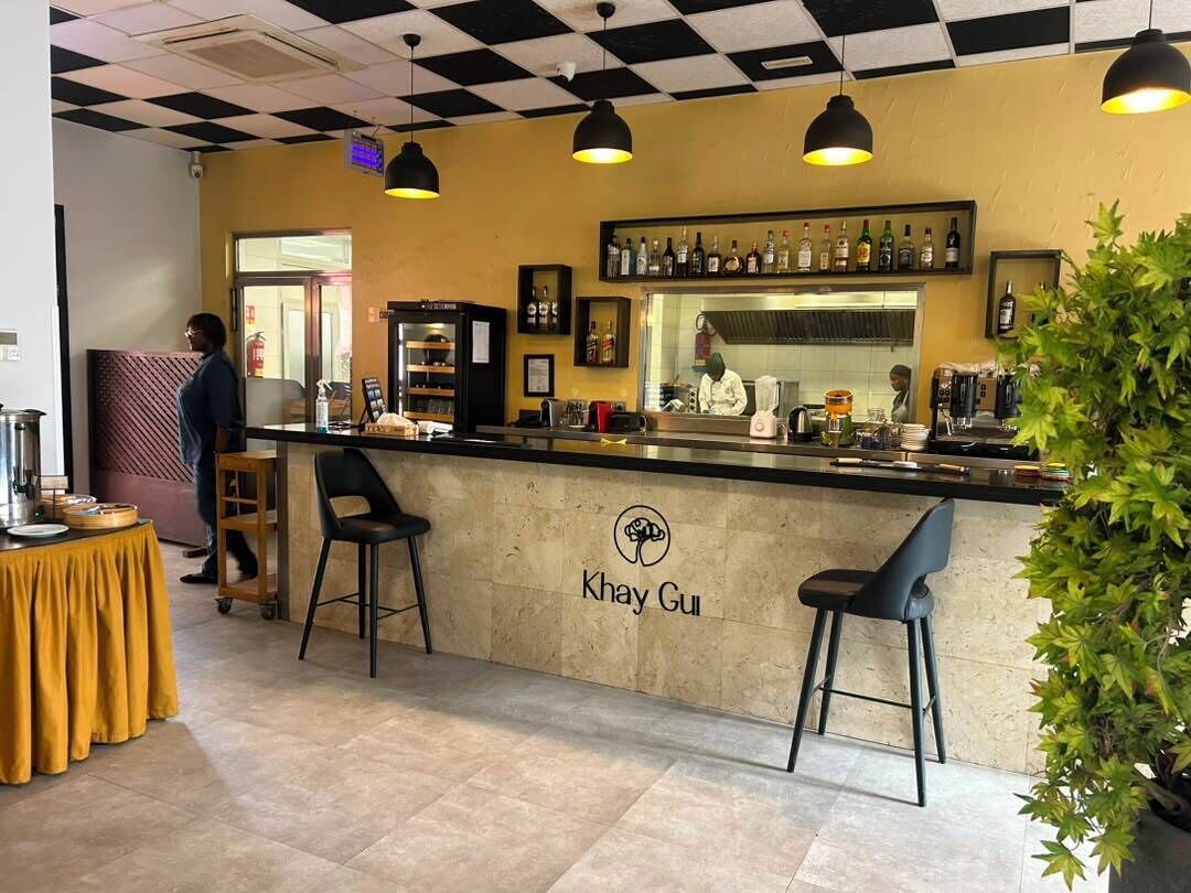 Interior de restaurante com teto xadrez, balcão de bar com banquetas, cozinha e área de refeições com toalha de mesa amarela.