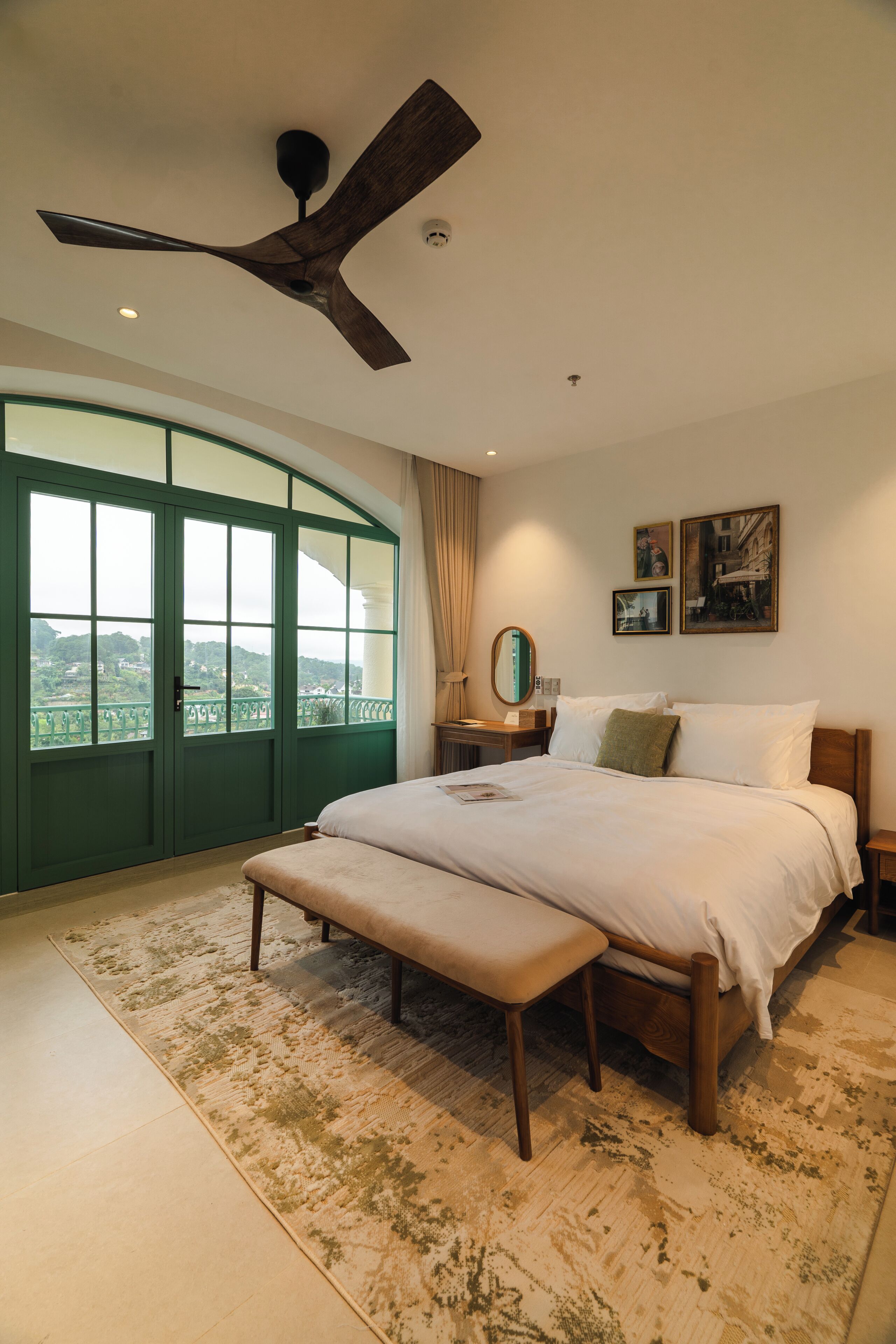 Quarto com uma cama grande, um banco, um ventilador de teto, vista por uma porta verde e uma obra de arte na parede.