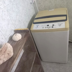Máquina de lavar com painel de controle, um banco de madeira e uma concha branca no chão.