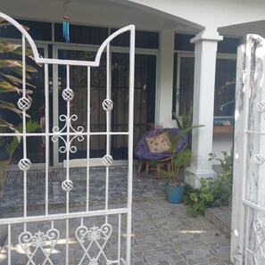 Uma entrada fechada por grades, com um portão de metal decorativo, um vaso de planta e uma cadeira de vime em frente a um edifício com janelas amplas.
