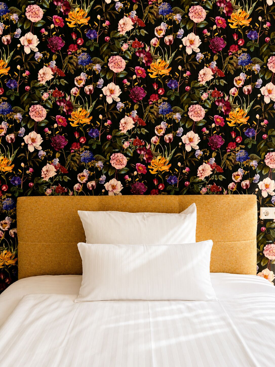 Cama com roupa de cama branca e duas almofadas, em frente a uma parede com papel de parede estampado com flores.