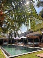 Uma área de piscina com espreguiçadeiras e guarda-sóis, cercada por palmeiras e uma estrutura com telhado de palha.