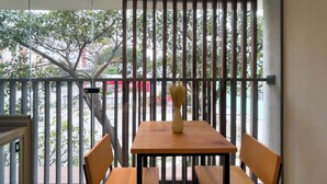 Uma varanda com mesa e cadeiras, um vaso com plantas secas, e vista para árvores e edifícios.