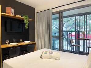 Quarto de hotel com cama, uma escrivaninha com duas cadeiras, televisão na parede e varanda com cadeira e guarda-corrimão.