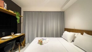 Quarto de hotel com cama, escrivaninha com cadeira, televisão e janela com cortinas.