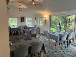 Uma área de refeições com várias mesas e cadeiras, grandes janelas com vista para o verde e teto com vigas aparentes e luminárias pendentes.
