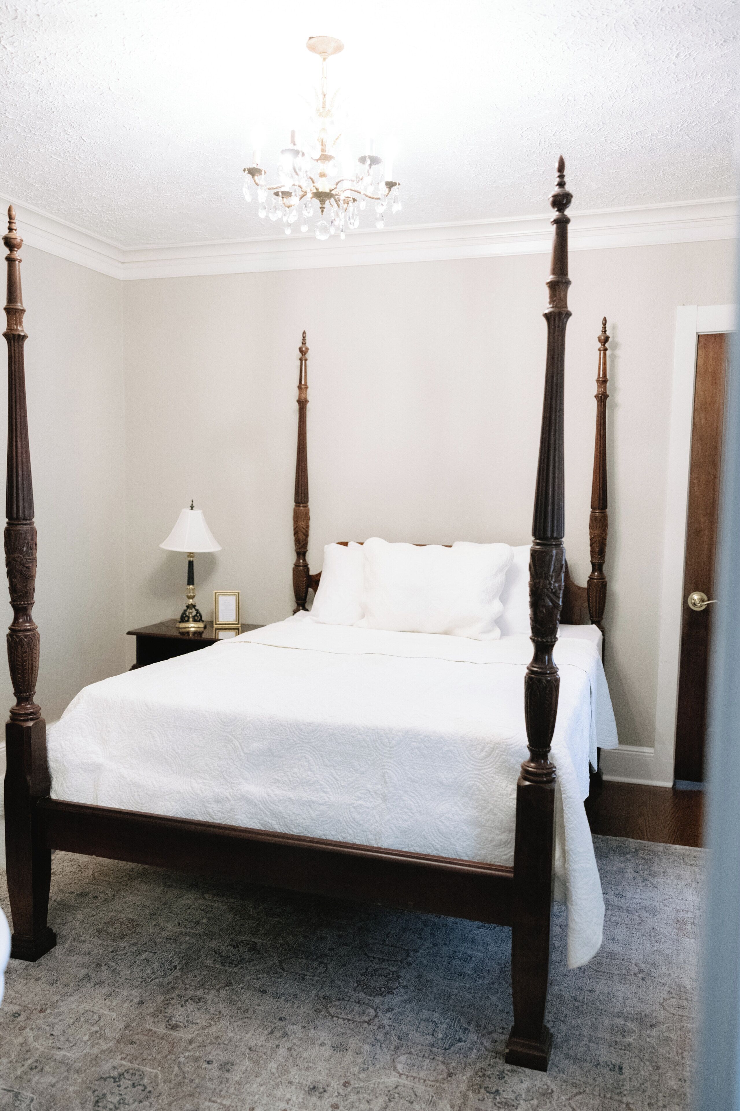 Uma cama com dossel, um candelabro, uma mesa de cabeceira com abajur e um quadro emoldurado.
