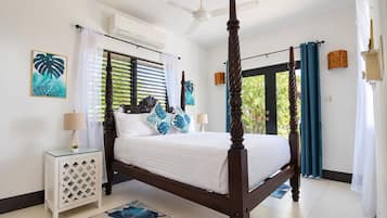 Uma cama de dossel com estrutura de madeira, roupa de cama branca e almofadas com padrão azul e branco. Há uma mesinha de cabeceira branca com uma luminária, um tapete floral azul e uma janela com persianas.