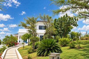 Uma casa moderna de dois andares com uma entrada em curvas, cercada por vegetação exuberante e um céu azul límpido.