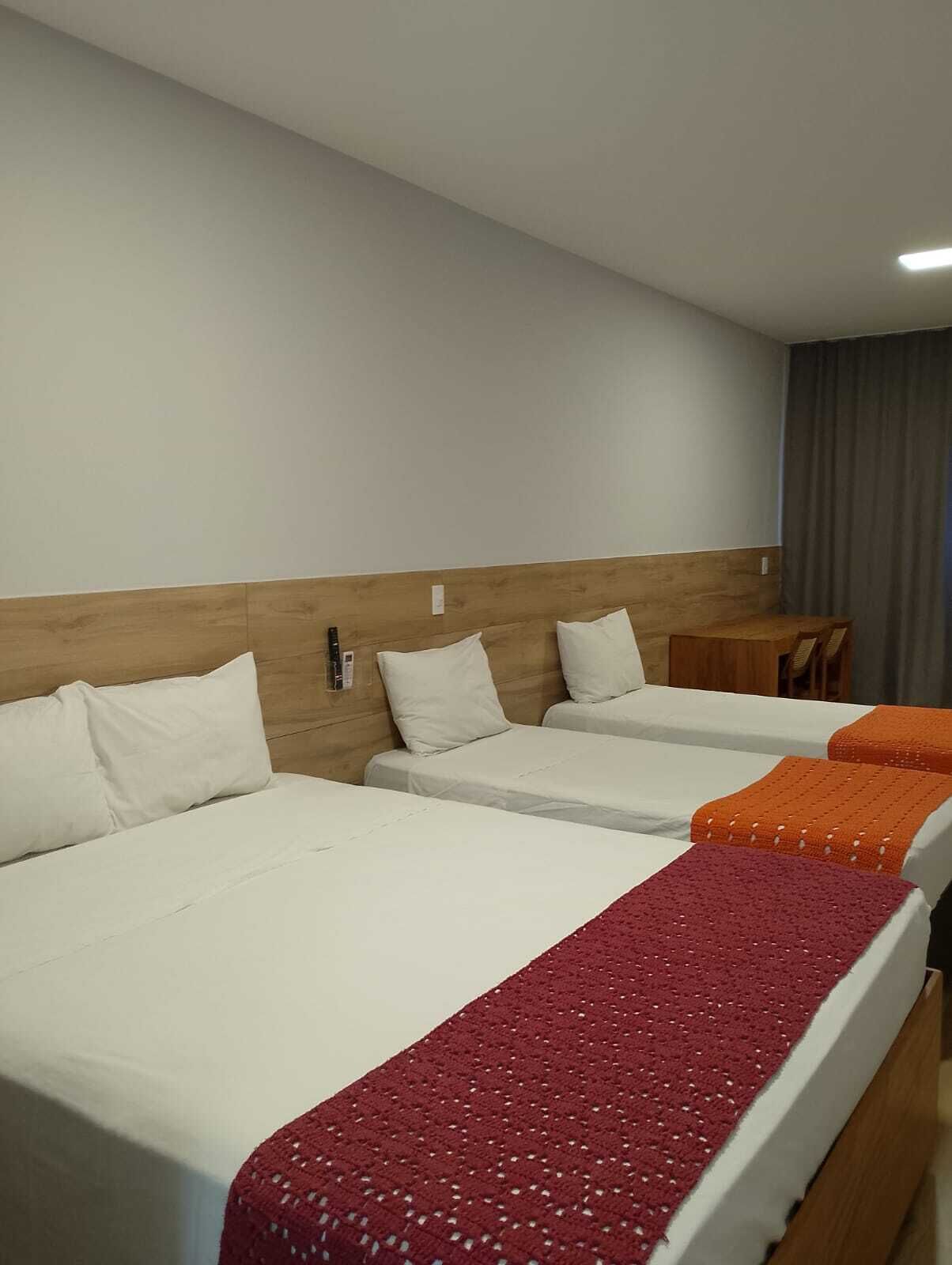Quarto de hotel com três camas, cabeceira de madeira, uma mesinha e uma cortina.