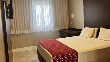 Quarto de hotel com cama, escrivaninha, televisão e janela com cortinas.