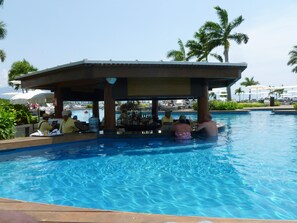 Uma área de bar à beira da piscina, com piscina, palmeiras e espreguiçadeiras.