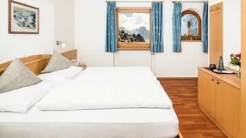 Quarto de hotel com cama, mesa de cabeceira, janela com cortinas, radiador, chão de madeira, guarda-roupa de madeira e um quadro na parede.