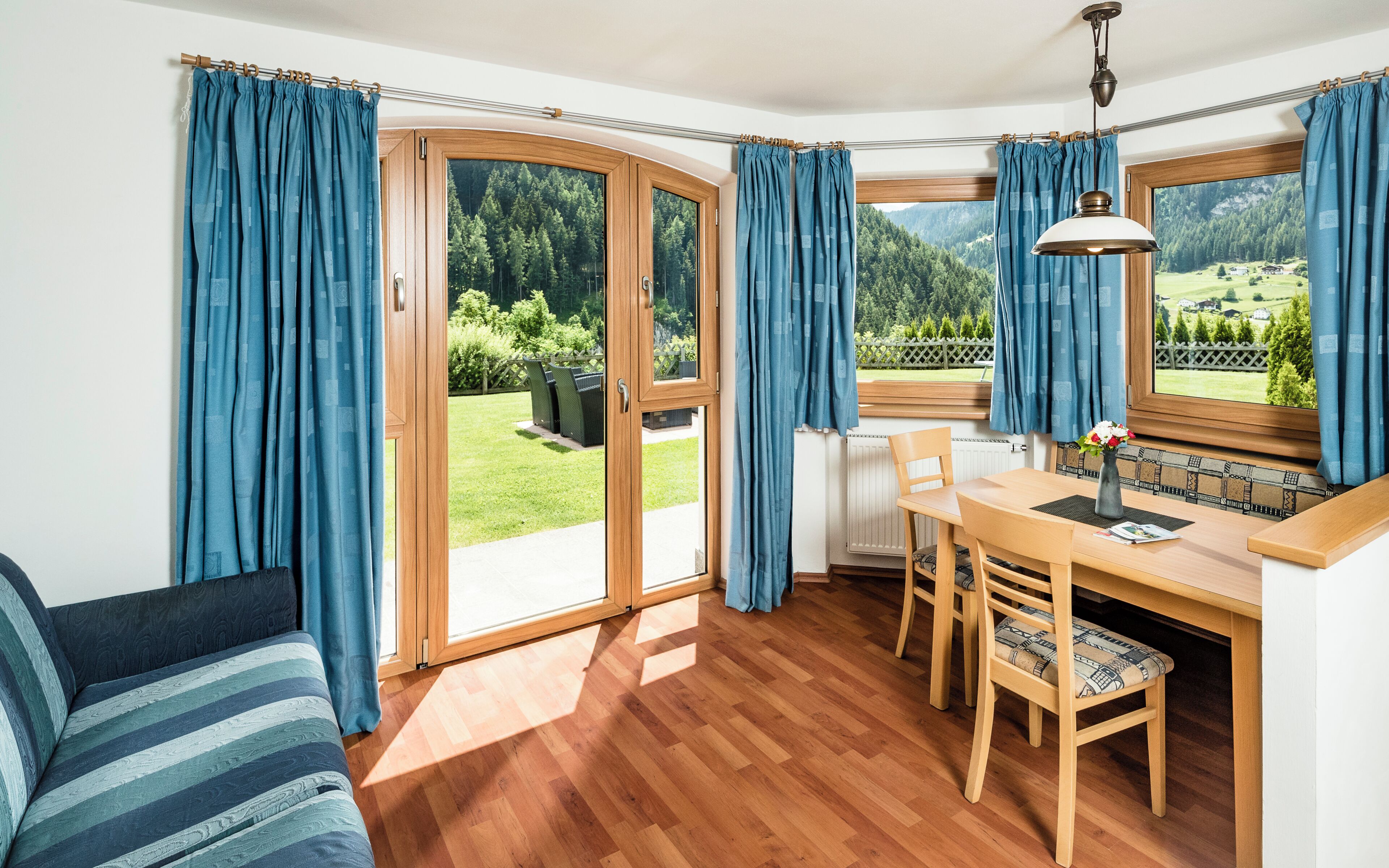 Quarto com piso de madeira, uma mesa de jantar com cadeiras, um sofá e vista para uma paisagem verde através de grandes janelas.