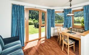 Quarto com piso de madeira, uma mesa de jantar com cadeiras, um sofá e vista para uma paisagem verde através de grandes janelas.
