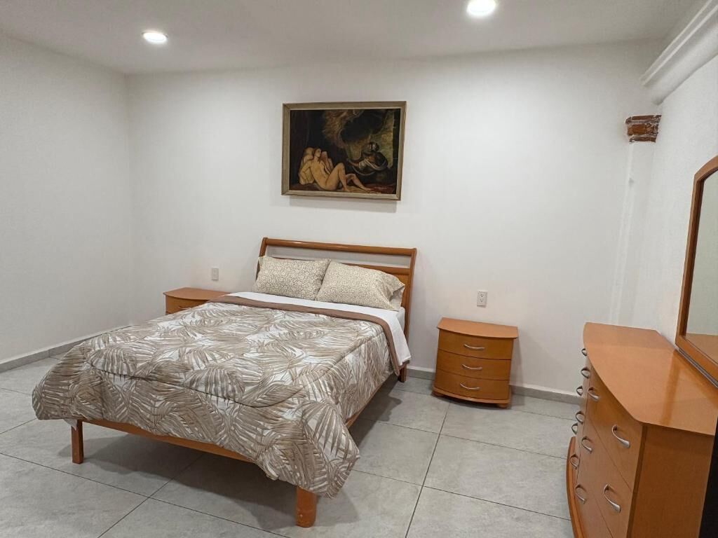 Quarto com uma cama, penteadeira de madeira, mesa de cabeceira, um quadro na parede e um espelho.