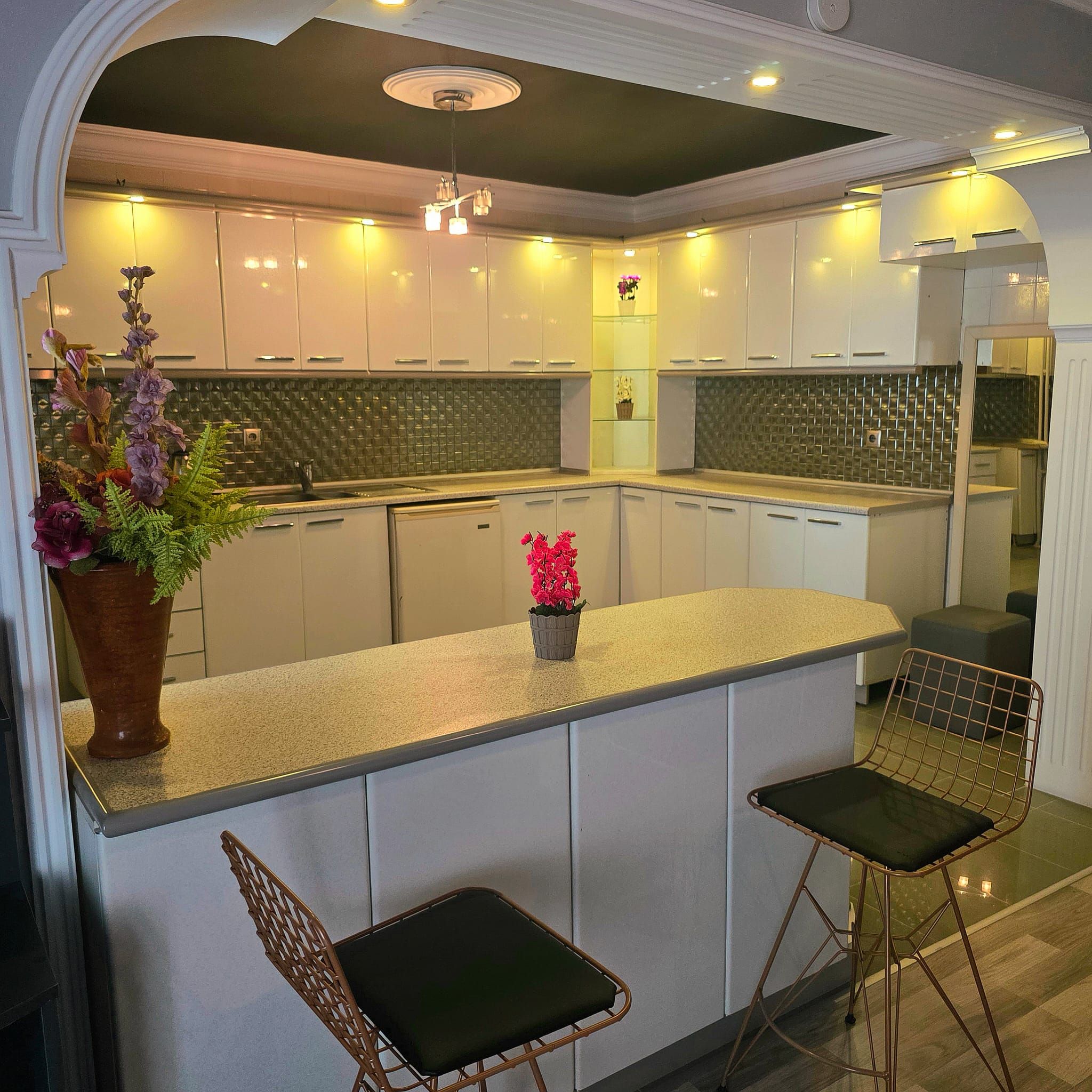 Cozinha moderna com armários brancos, uma ilha central com dois bancos e uma planta em vaso.