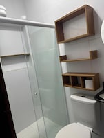 Casa de banho com cabine de chuveiro em vidro, sanita e prateleiras de madeira sobre a pia.