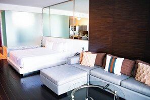 Quarto de hotel moderno com uma cama grande, um sofá, uma mesa de centro de vidro e vista para uma cozinha e banheiro através de um espelho amplo.
