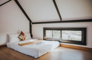Quarto com cama, travesseiros, janela e piso de madeira.