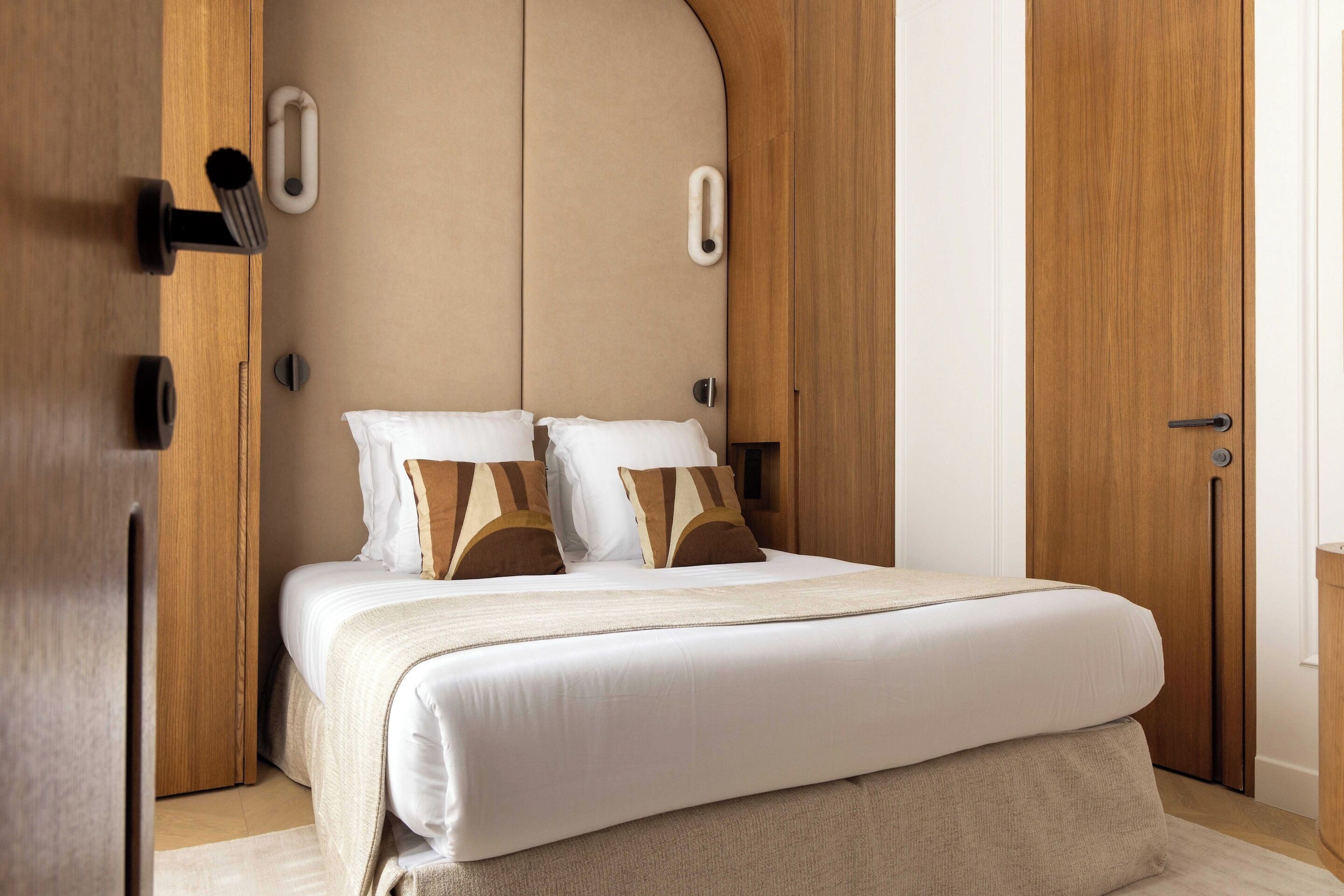 Cama bem arrumada com cabeceira, guarda-roupa de madeira e porta com puxador.