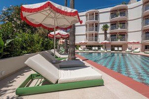 Uma área à beira da piscina com espreguiçadeiras e guarda-sóis, uma piscina e um prédio com varandas.